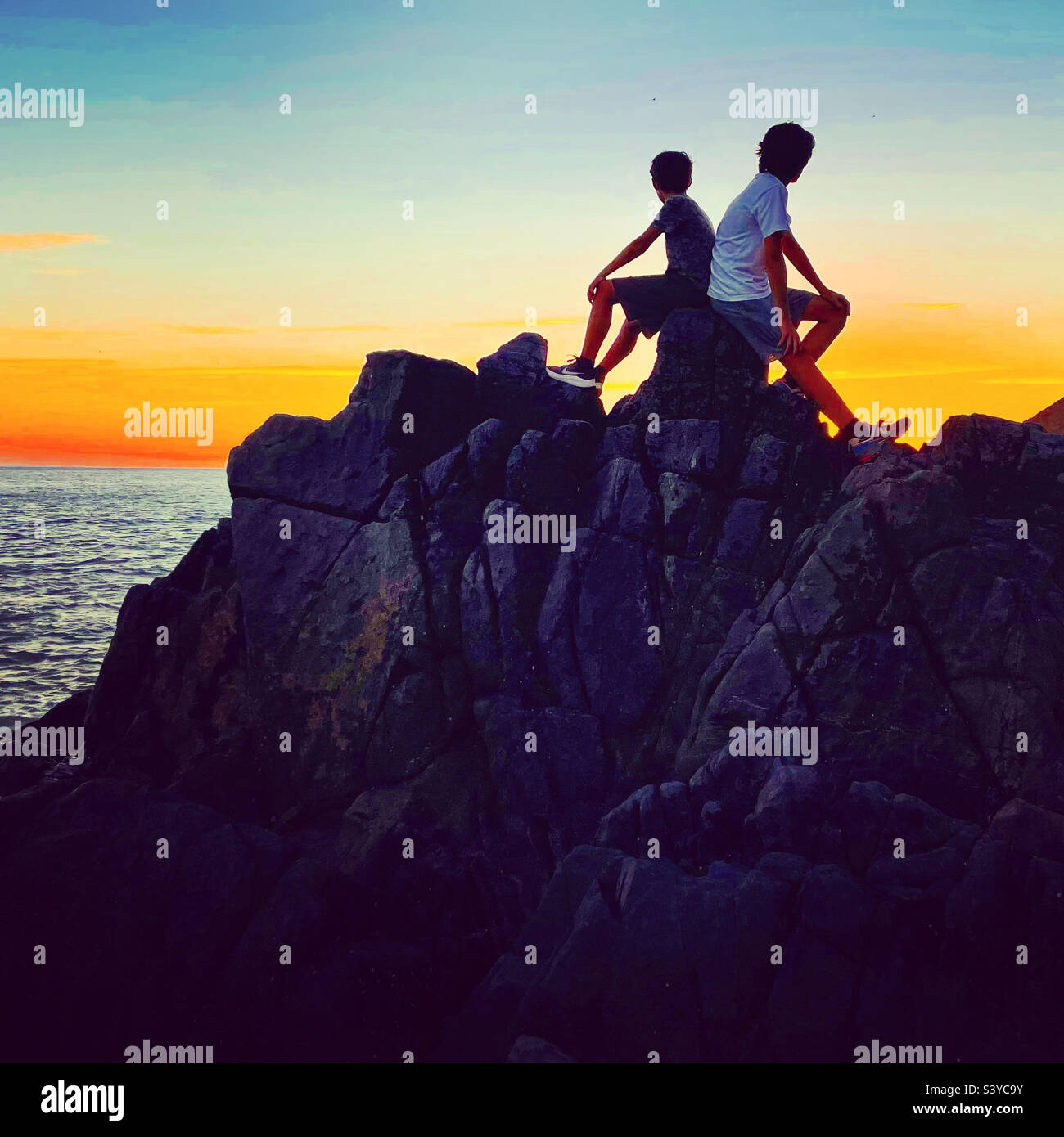 2 people sitting on a rick are watching the sunset at the ocean Stock ...