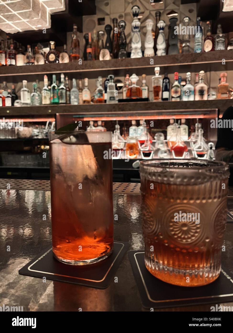 Two coctails and a bar on the background having colorful bottles of ...