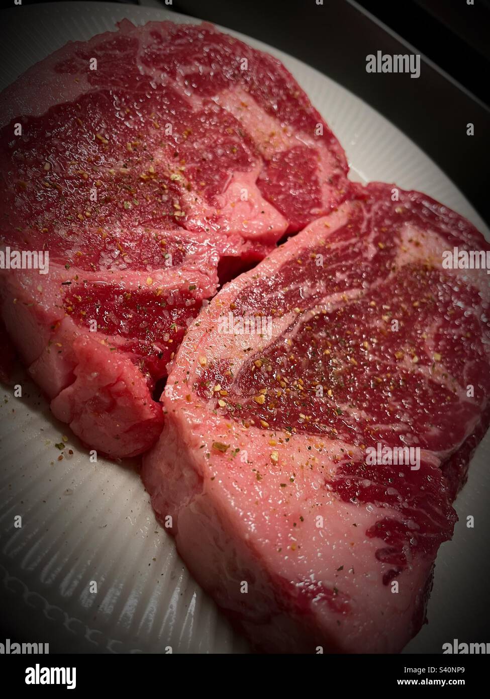 Close up of two raw ribeye steaks seasoned with salt and pepper await ...