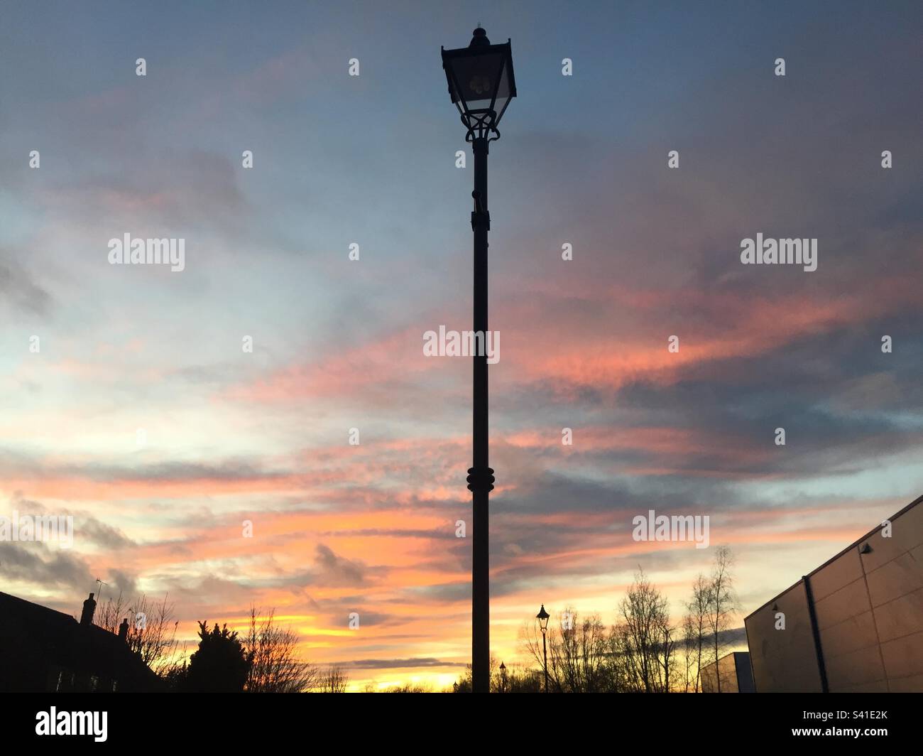 Sunset, Colourful sky with silhouetted lamp posts, trees and rooftops ...