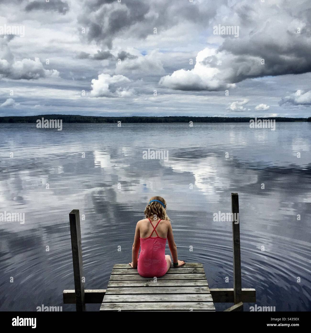 A young eight year old girl sits on a pier looking at dark storm clouds ...