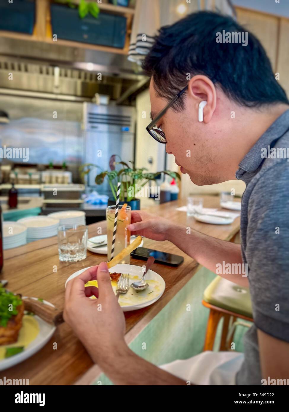 Asian man with wireless earbuds having a meal and a drink at bar ...