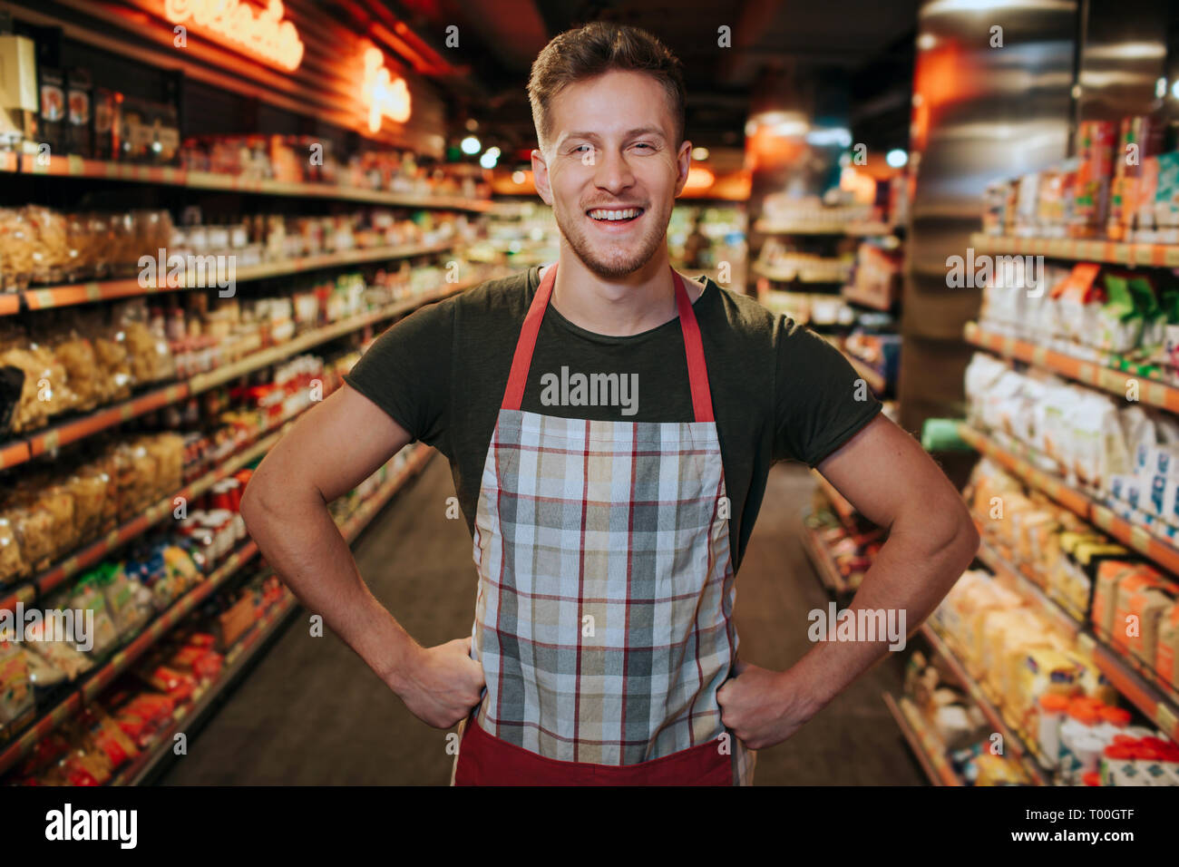 Happy cheerful positive young man in apron stand in grocery store among ...