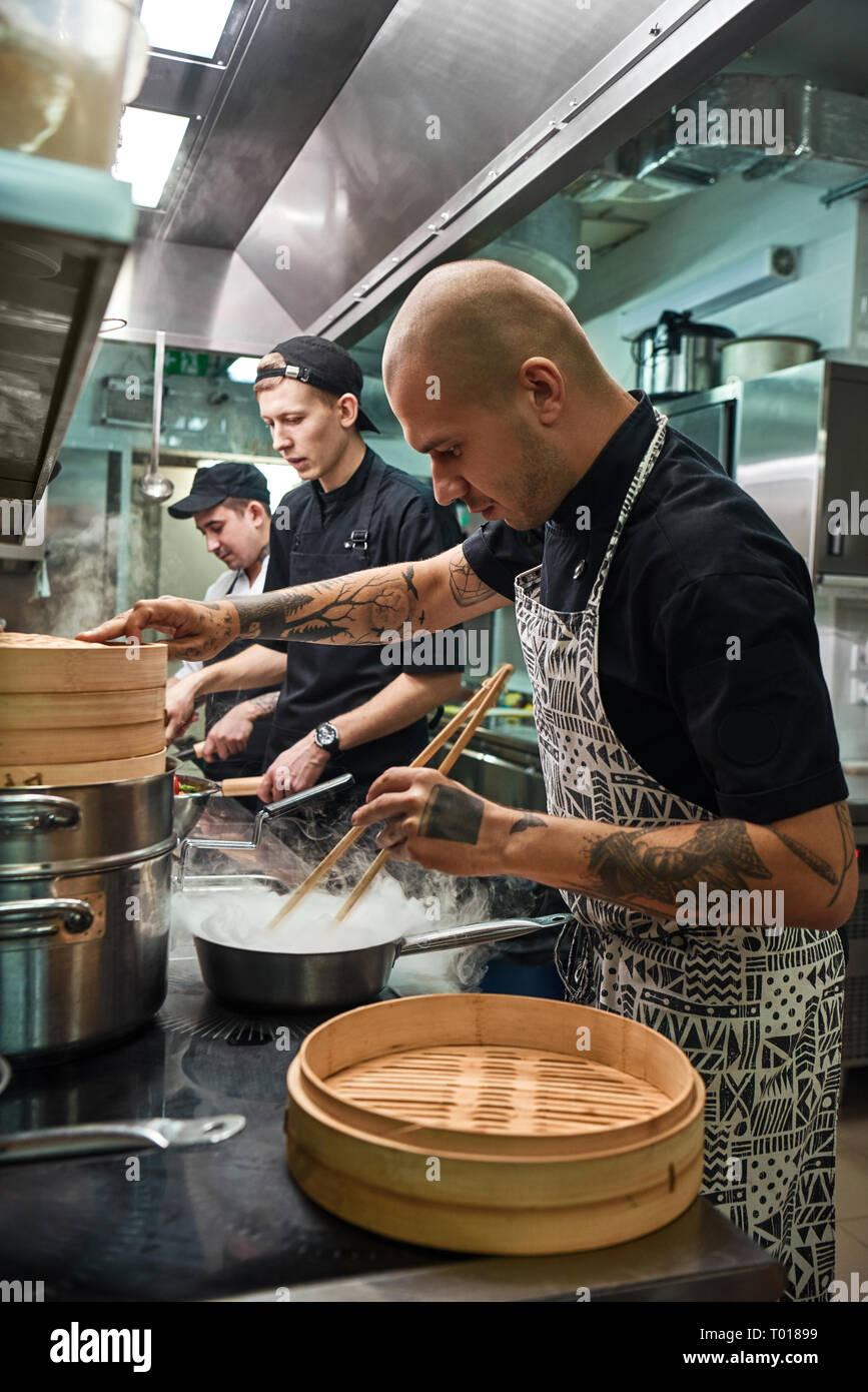 Full concentration. Young bald chef in apron with tattoos on his arms ...
