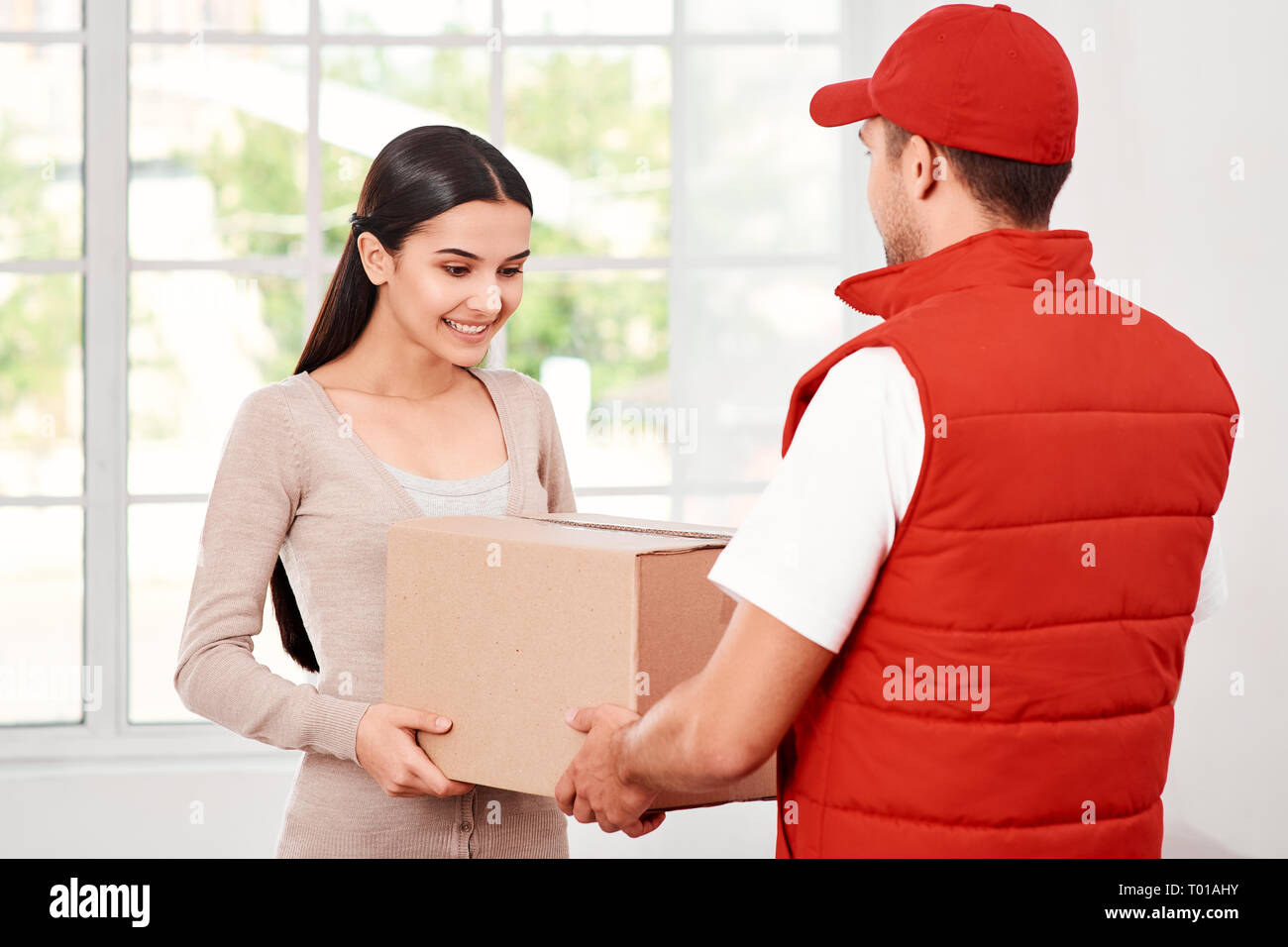 Cheerful postman wearing red postal uniform is delivering parcel to a ...