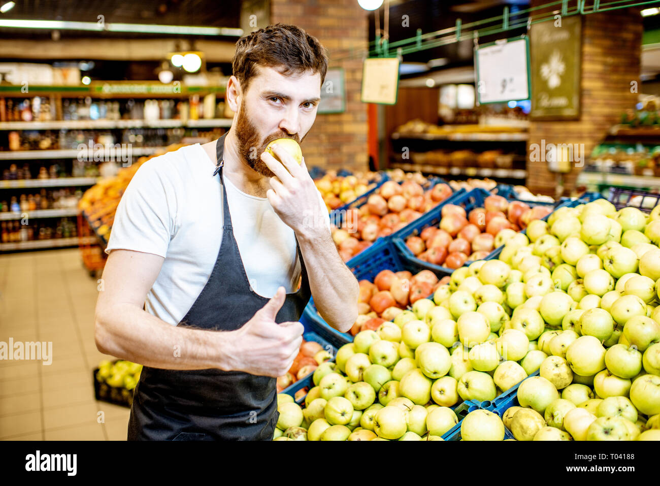 Handsome shop worker in uniform tasting apple checking the quality in ...