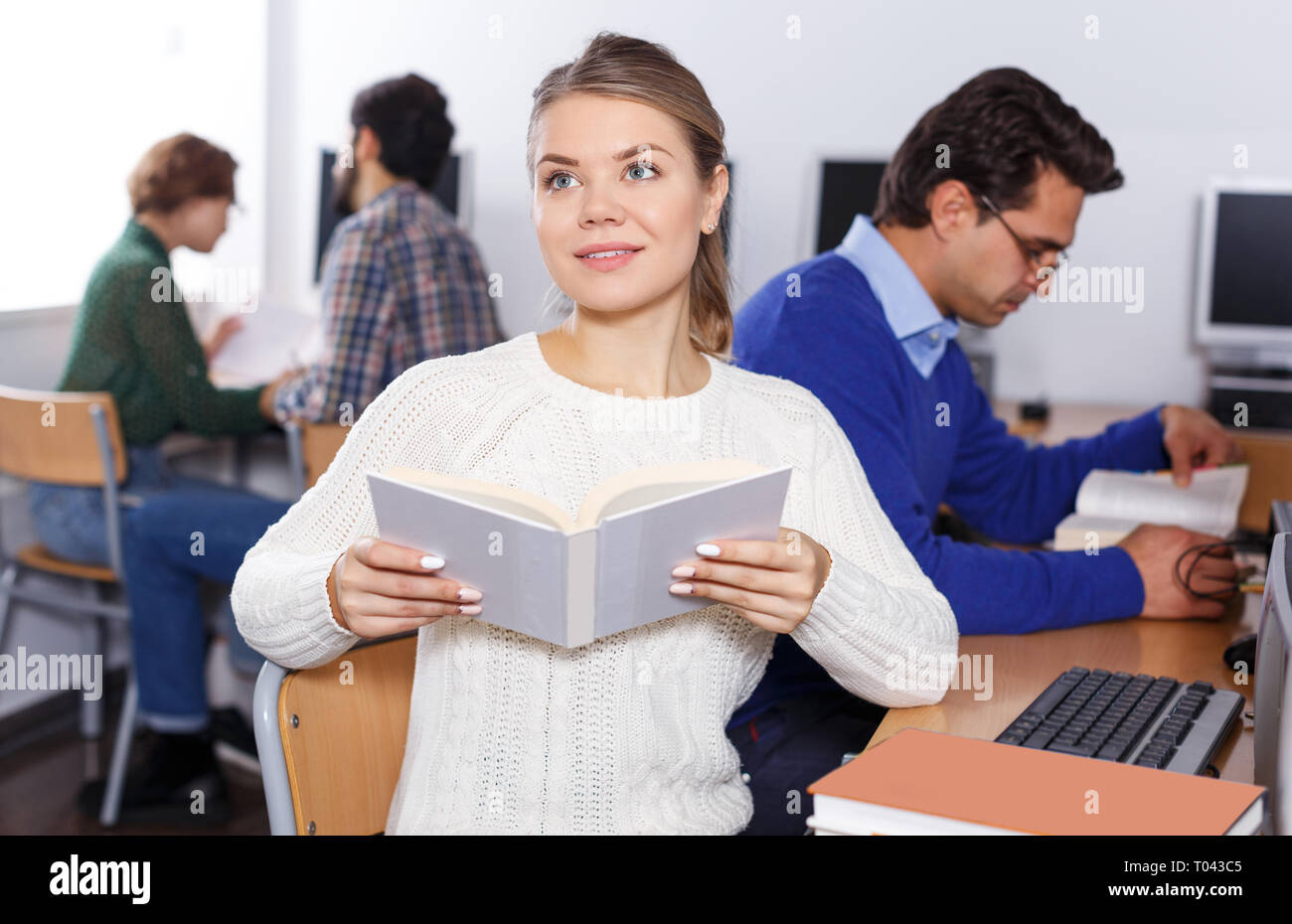 young female student reading textbook while studying in university ...