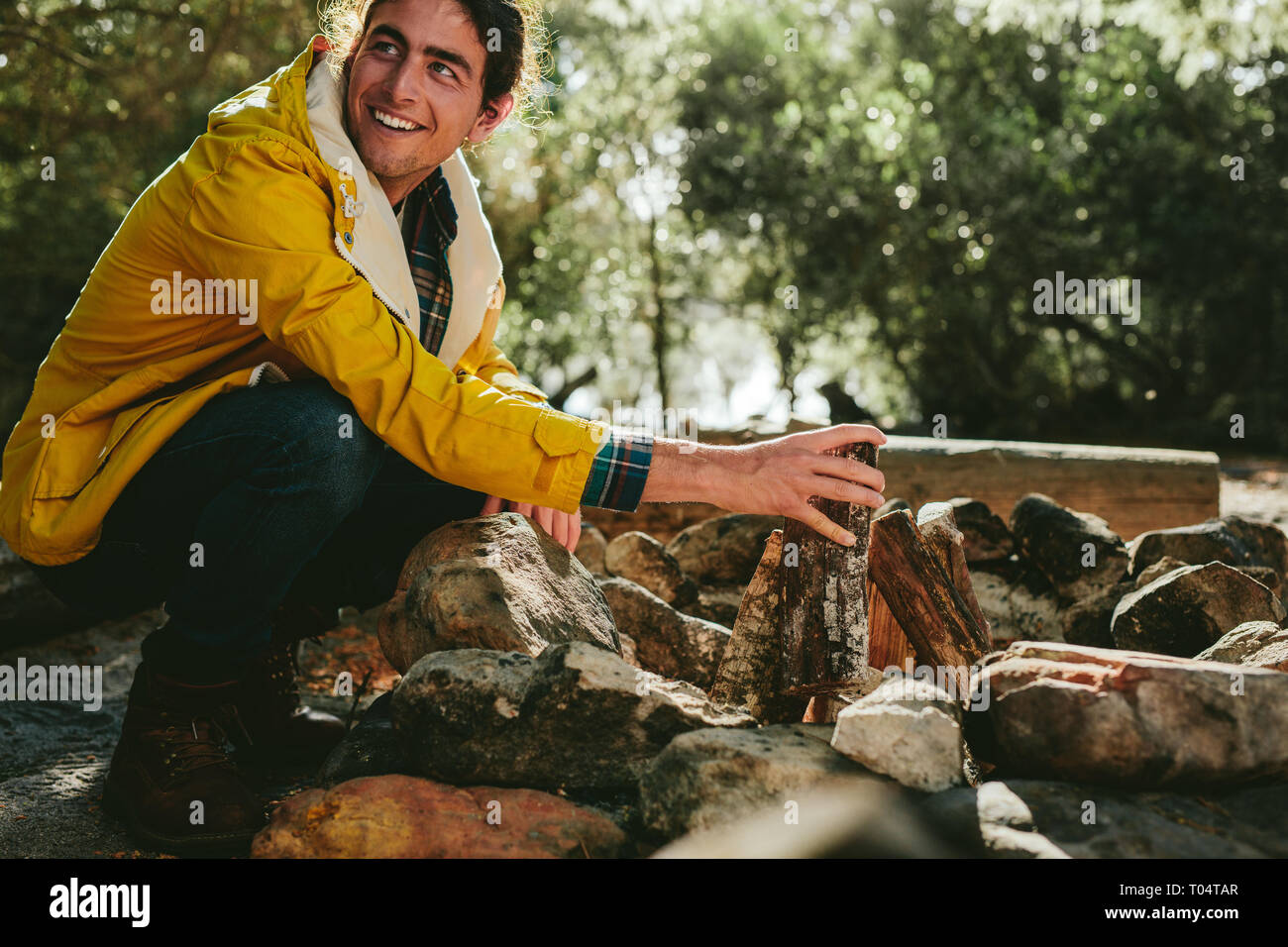 Smiling man putting fire wood setting up a bonfire in the countryside ...