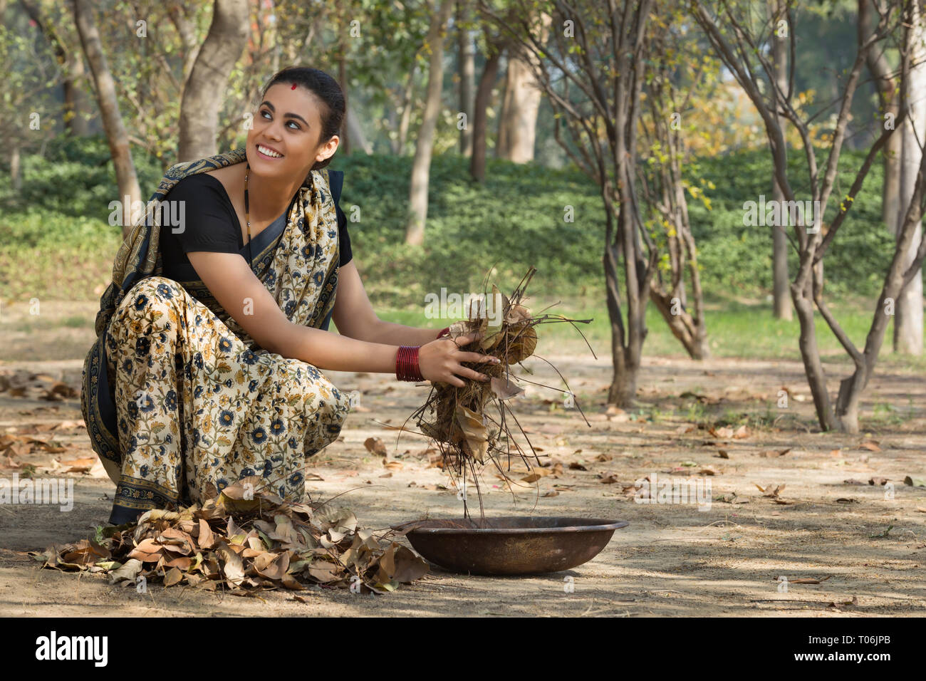 Smiling rural woman dressed in saree collecting dried leaves from ...