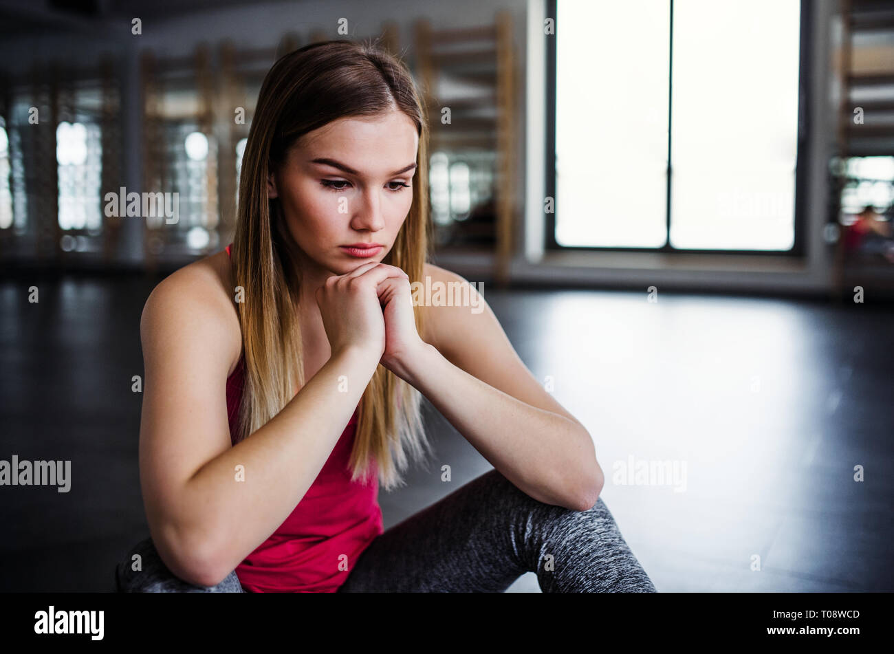A portrait of young sad and frustrated girl or woman sitting in a gym ...
