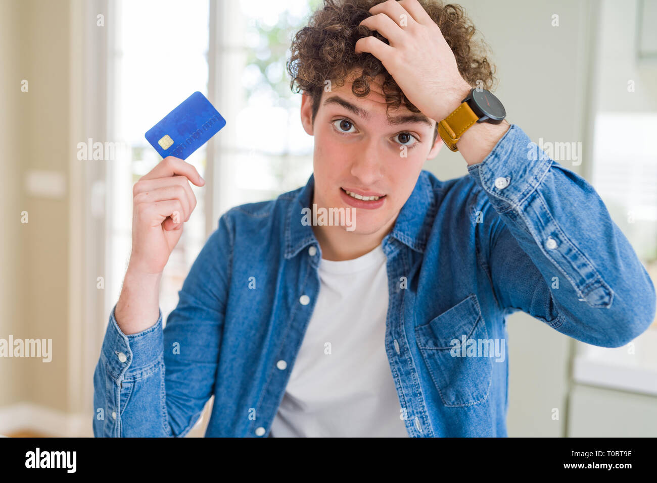 Young man holding credit card stressed with hand on head, shocked with ...