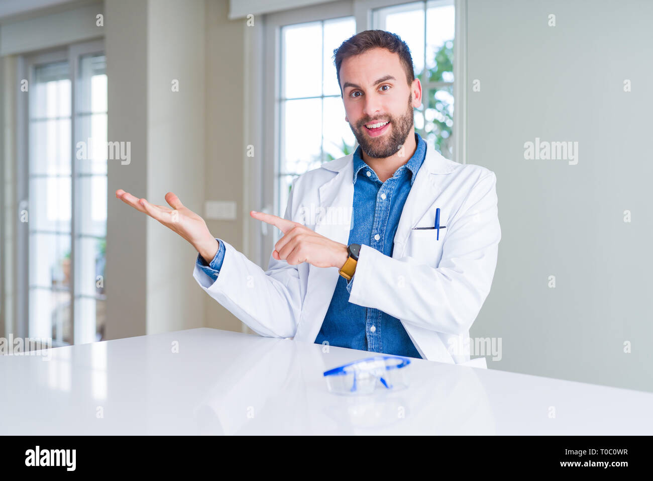 Handsome scientist man wearing white robe and safety glasses amazed and ...