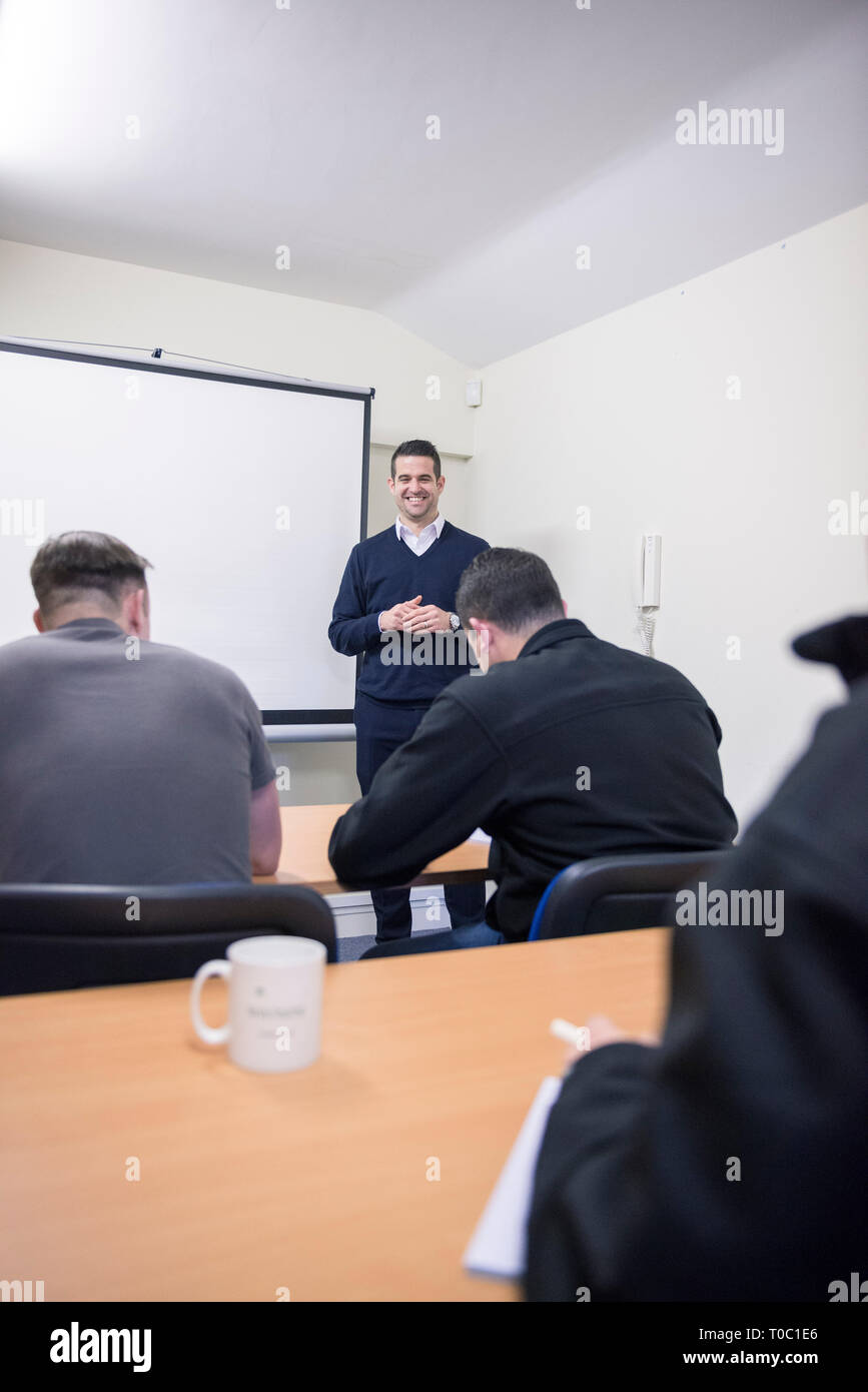 A white man in a blue jumper holds a training presentation for fellow ...