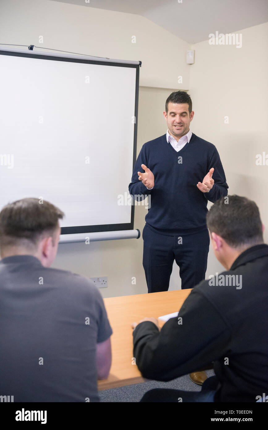A white man in a blue jumper holds a training presentation for fellow ...
