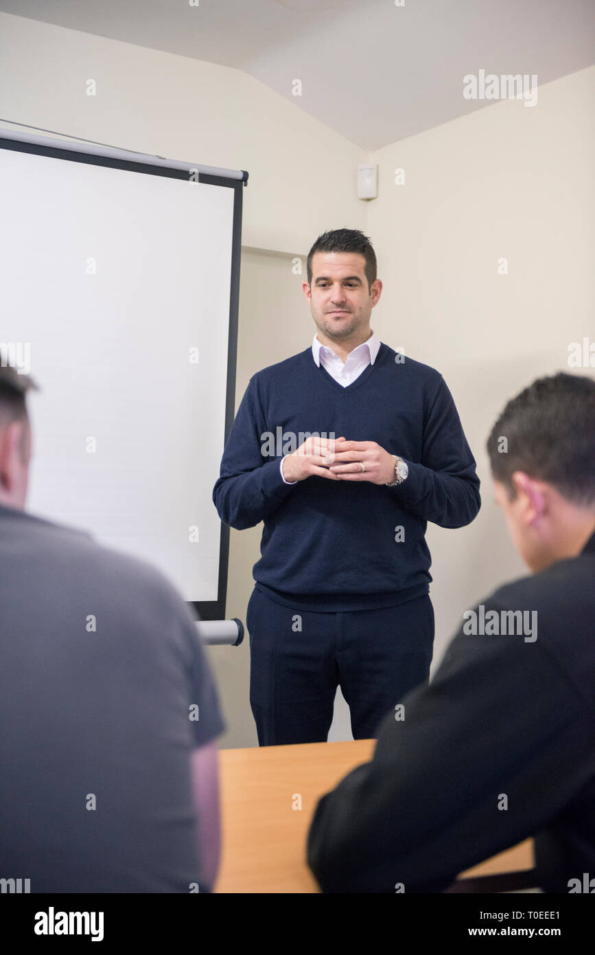 A white man in a blue jumper holds a training presentation for fellow ...