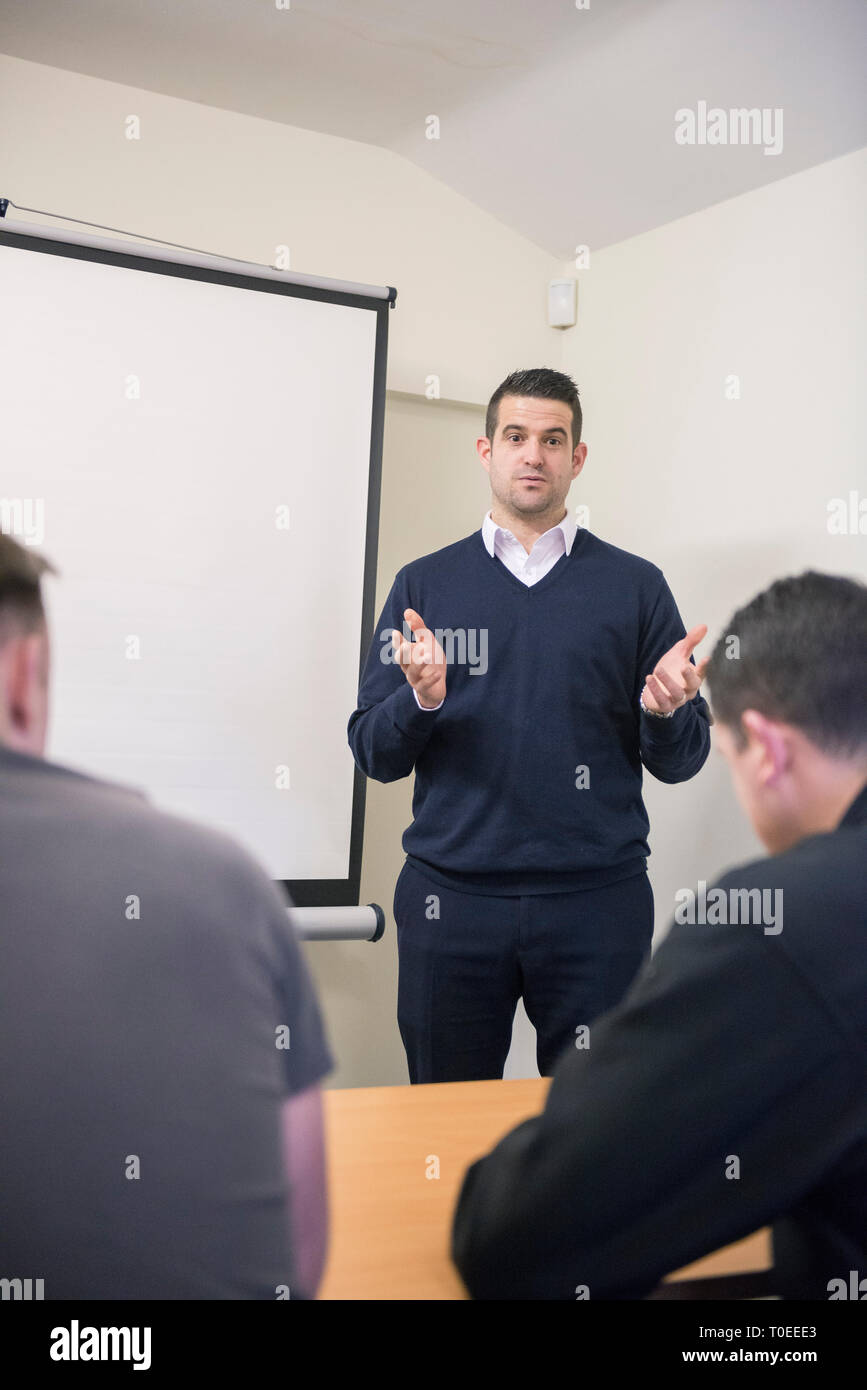 A white man in a blue jumper holds a training presentation for fellow ...