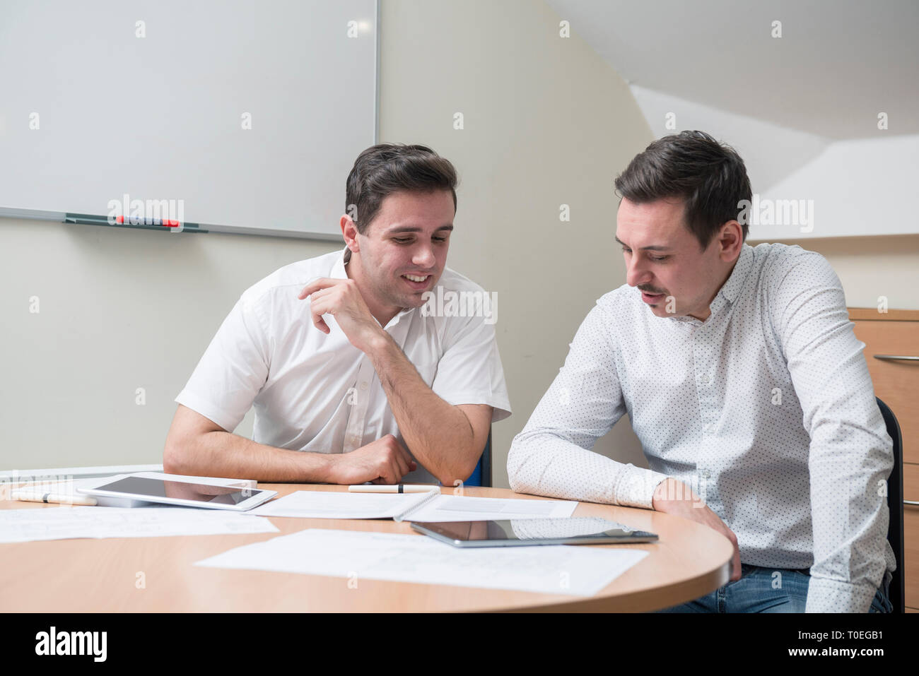 Two men sit round a table in a small office meeting room to review ...