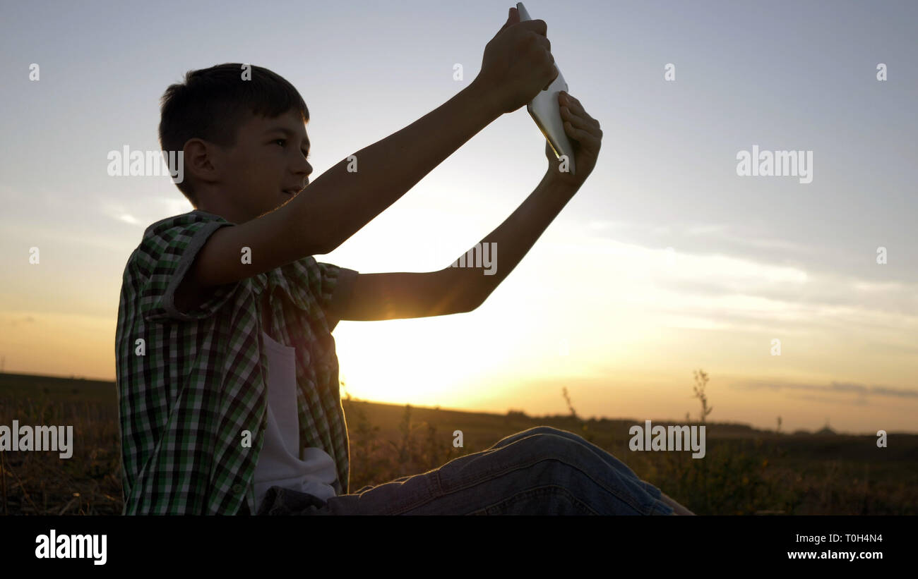 boy talking on video call on tablet sitting in field at sunset, have ...