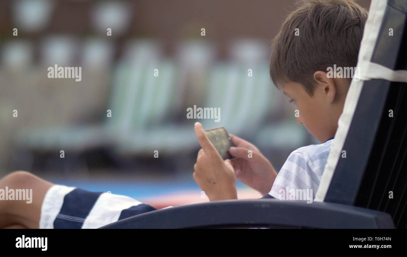 Caucasian teen boy lying on lounger operating mobile phone at swimming ...