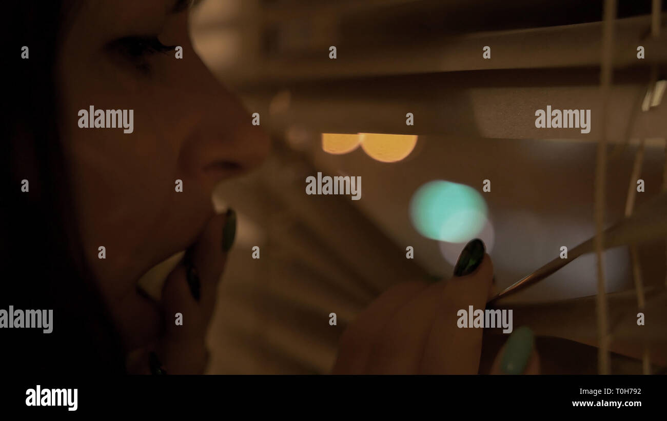 Cute brunette girl standing by the window at night, pushing the blinds ...