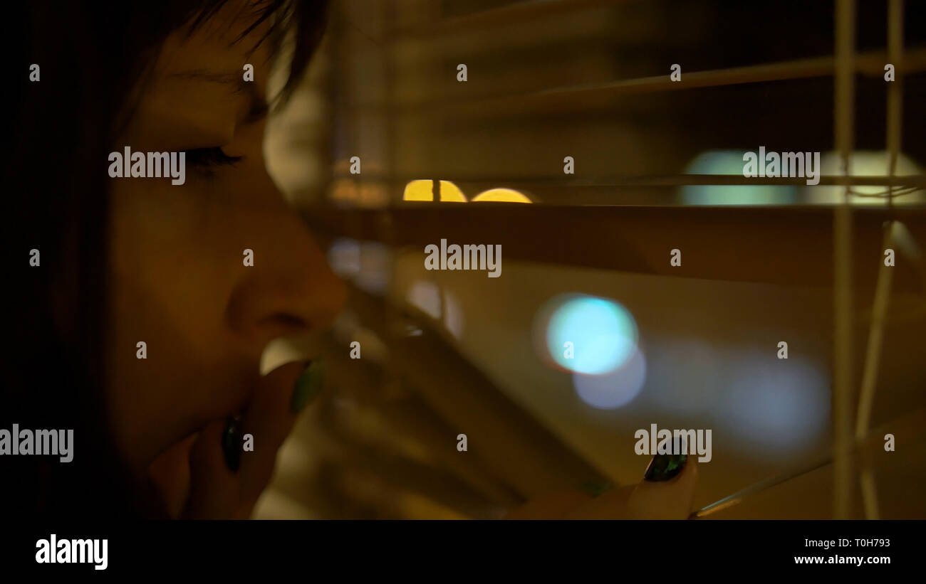 Cute brunette girl standing by the window at night, pushing the blinds ...