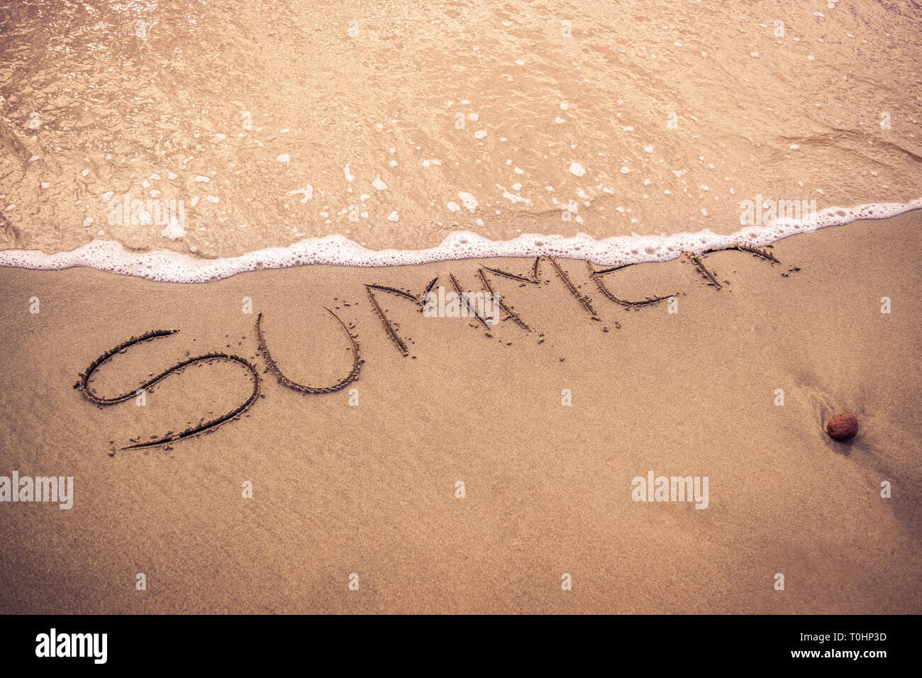 The word "summer" written on the sand by the sea at sunset while a wave ...