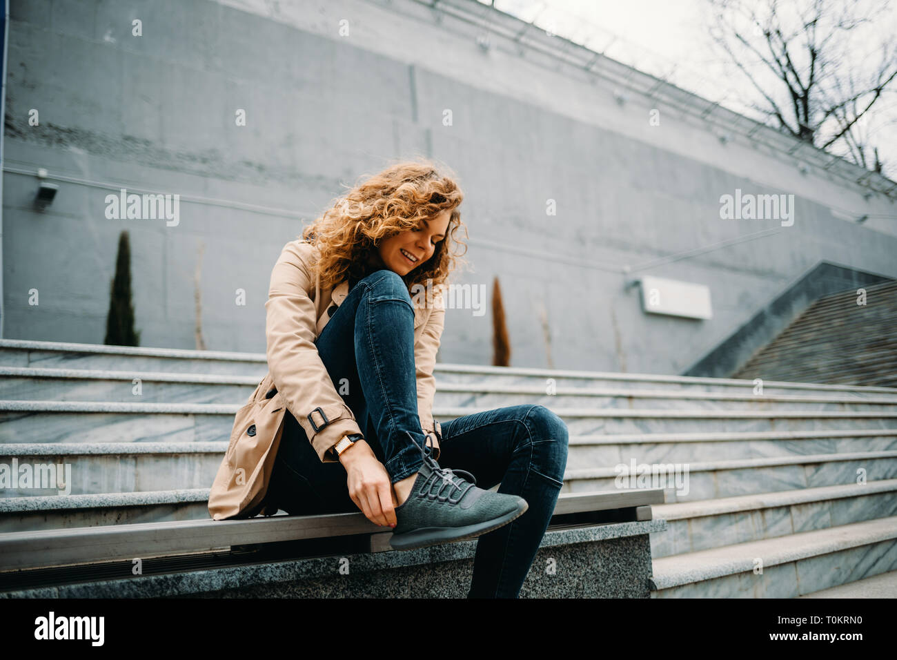Happy young woman wearing beige trench coat and blue jeans sitting on gray stone stairs ...