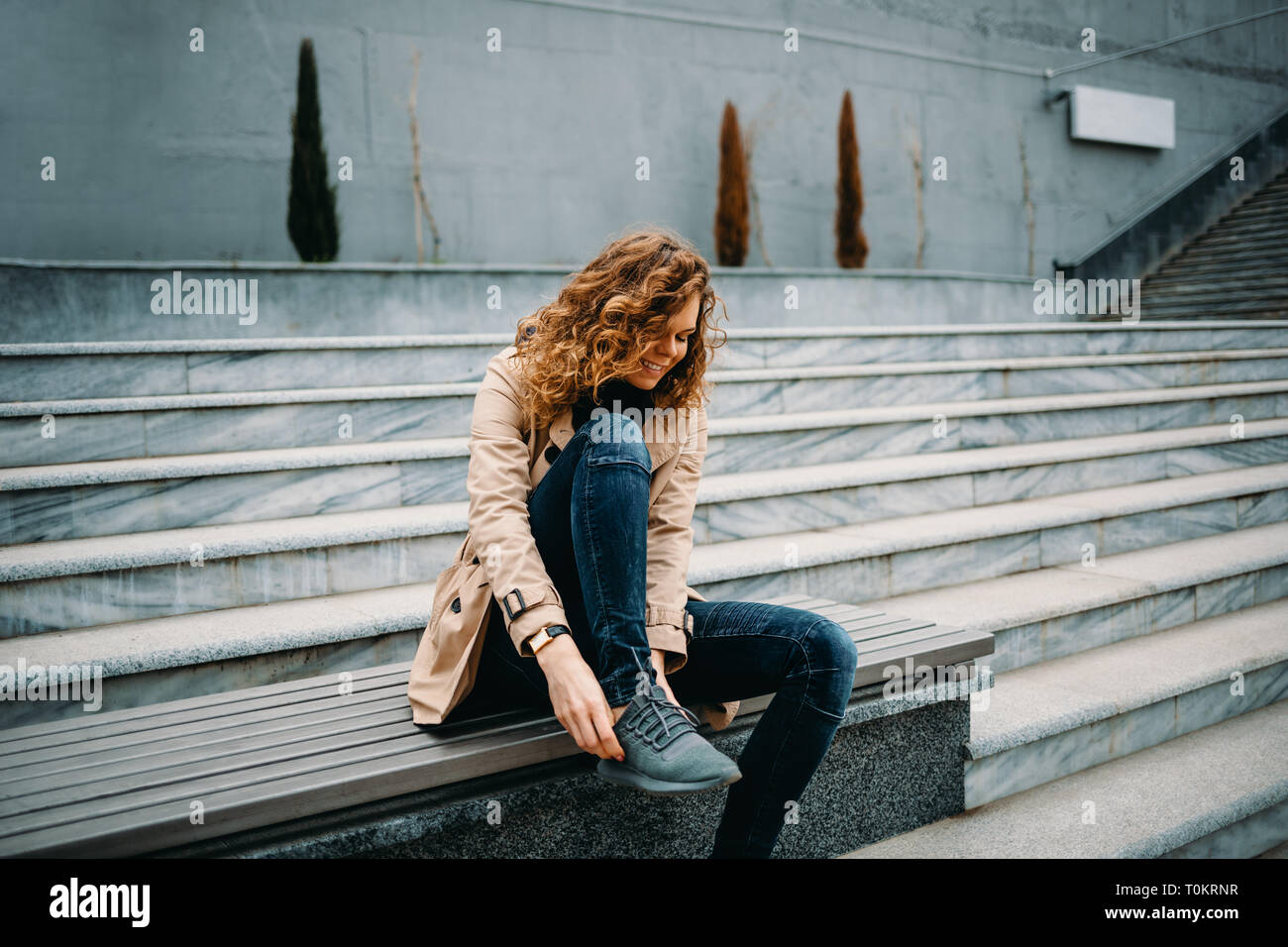 Smiling young woman wearing beige trench coat sitting on gray stone stairs adjusting her ...