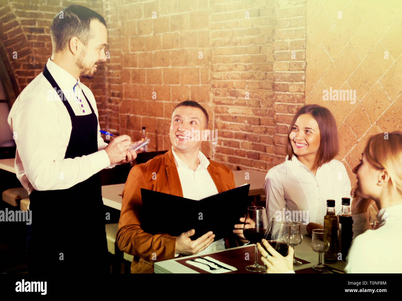 Cheerful young man waiter receiving order from guests in country ...