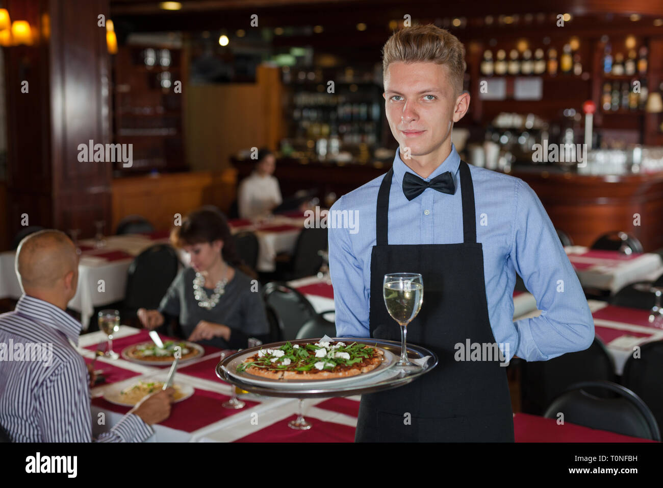 Polite waiter holding tray in restaurant with customers behind him ...
