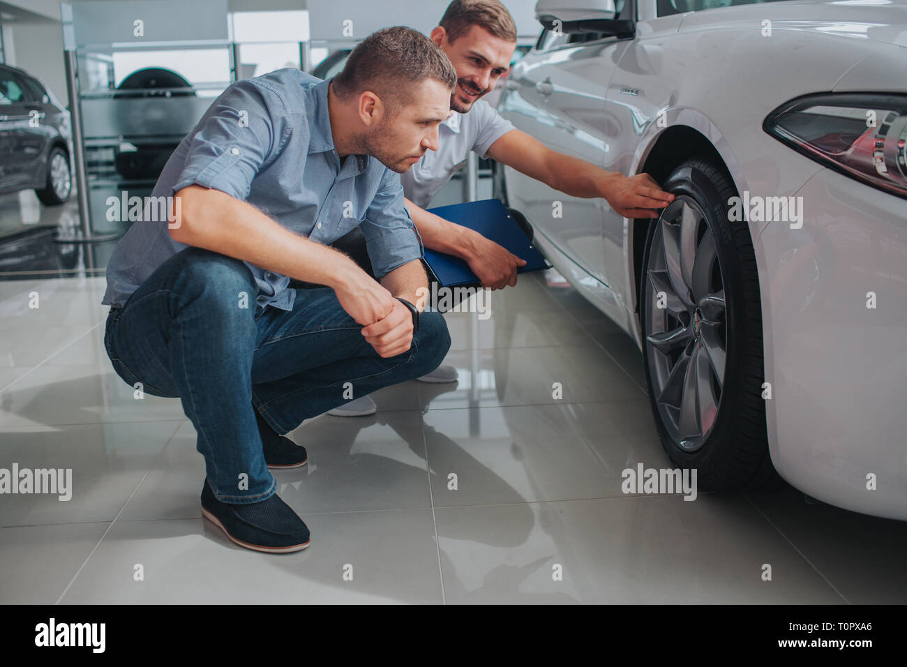 Two men sit in squad position and look at wheel of white car. Berded ...