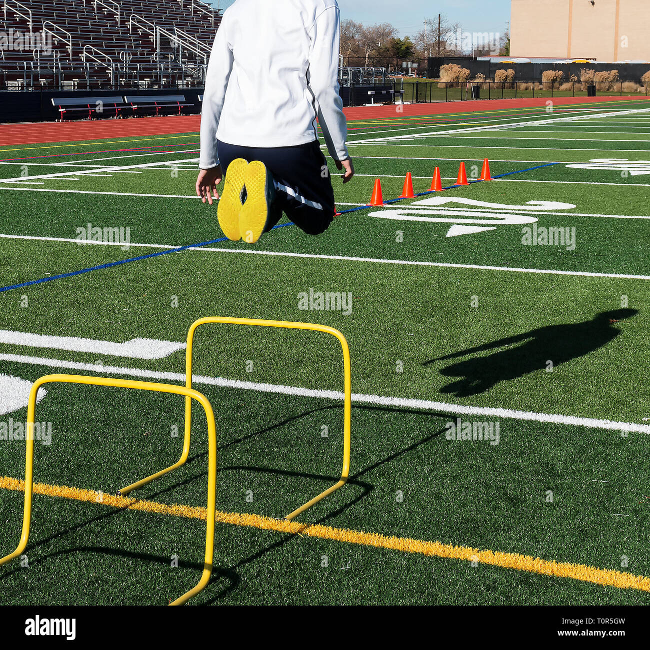 A high school track and field athlete jumps over yellow hurdles with orange cones in the ...