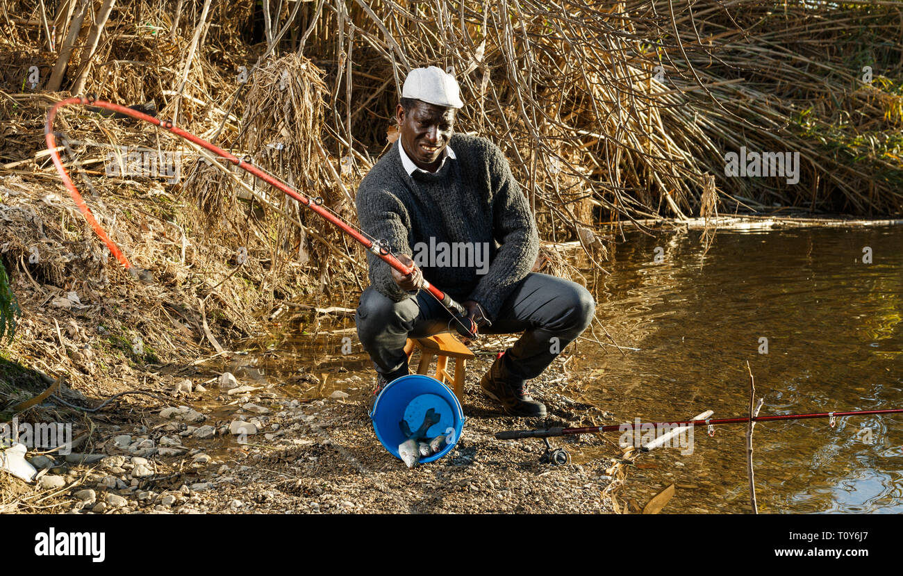 Portrait of enthusiastic African man pulling fish with rods on river ...