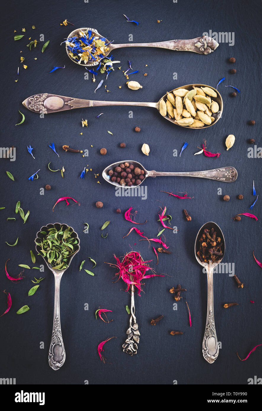 Silver spoons full of spices, herbs and floral teas, on black slate ...