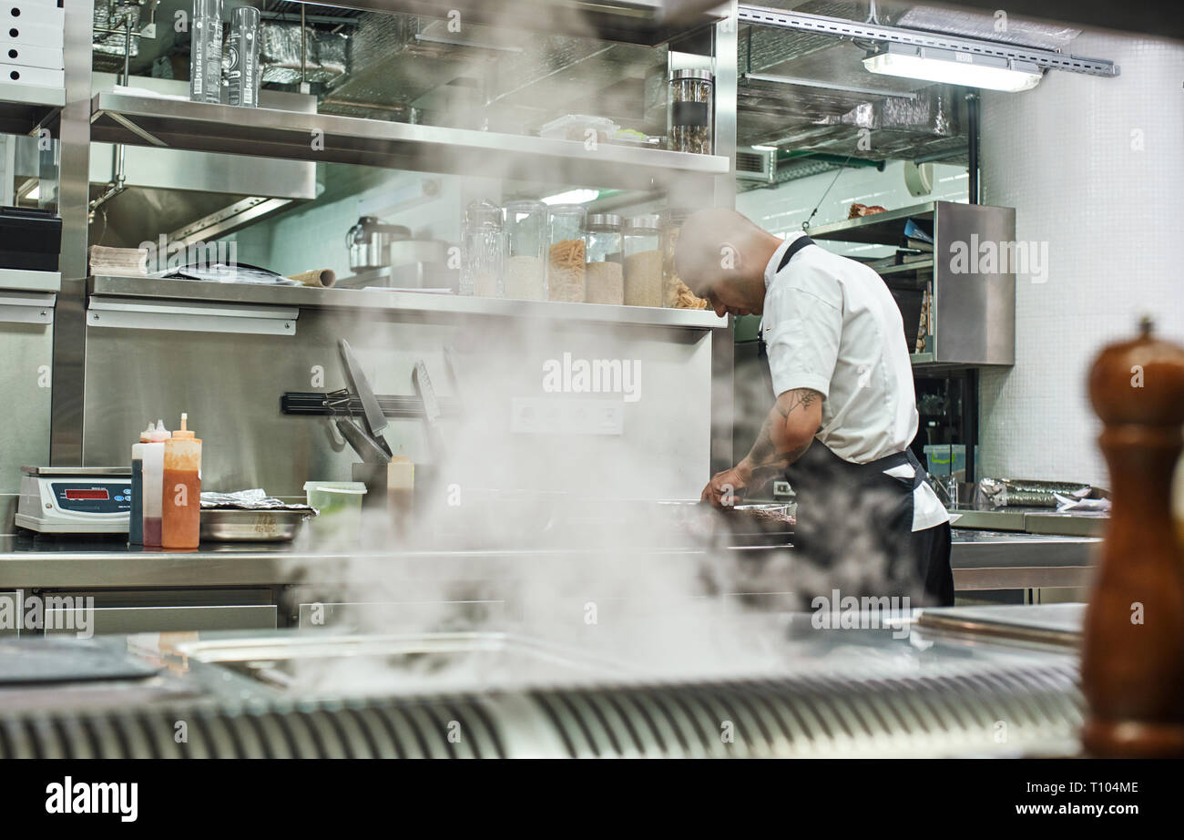 Busy day. Back view of male chef in apron cutting a meat while standing ...