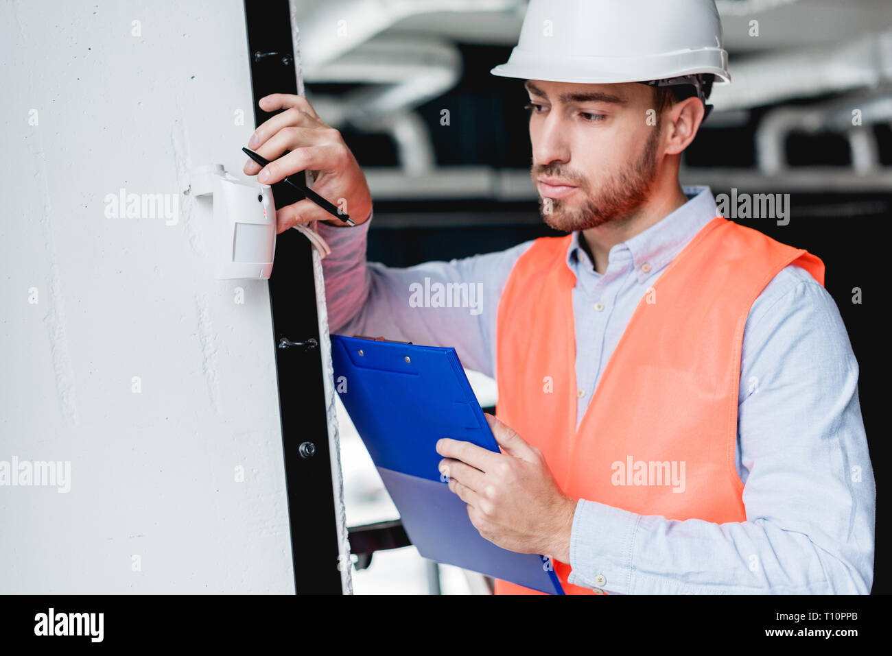 handsome fireman in helmet checking fire alarm while holding clipboard ...