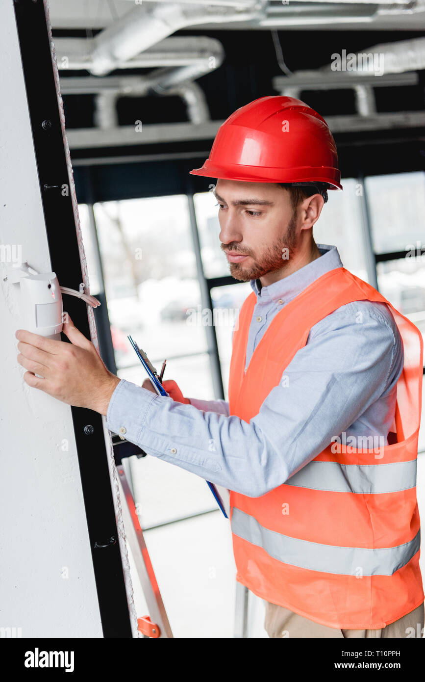 pensive fireman in helmet checking fire alarm while holding clipboard ...