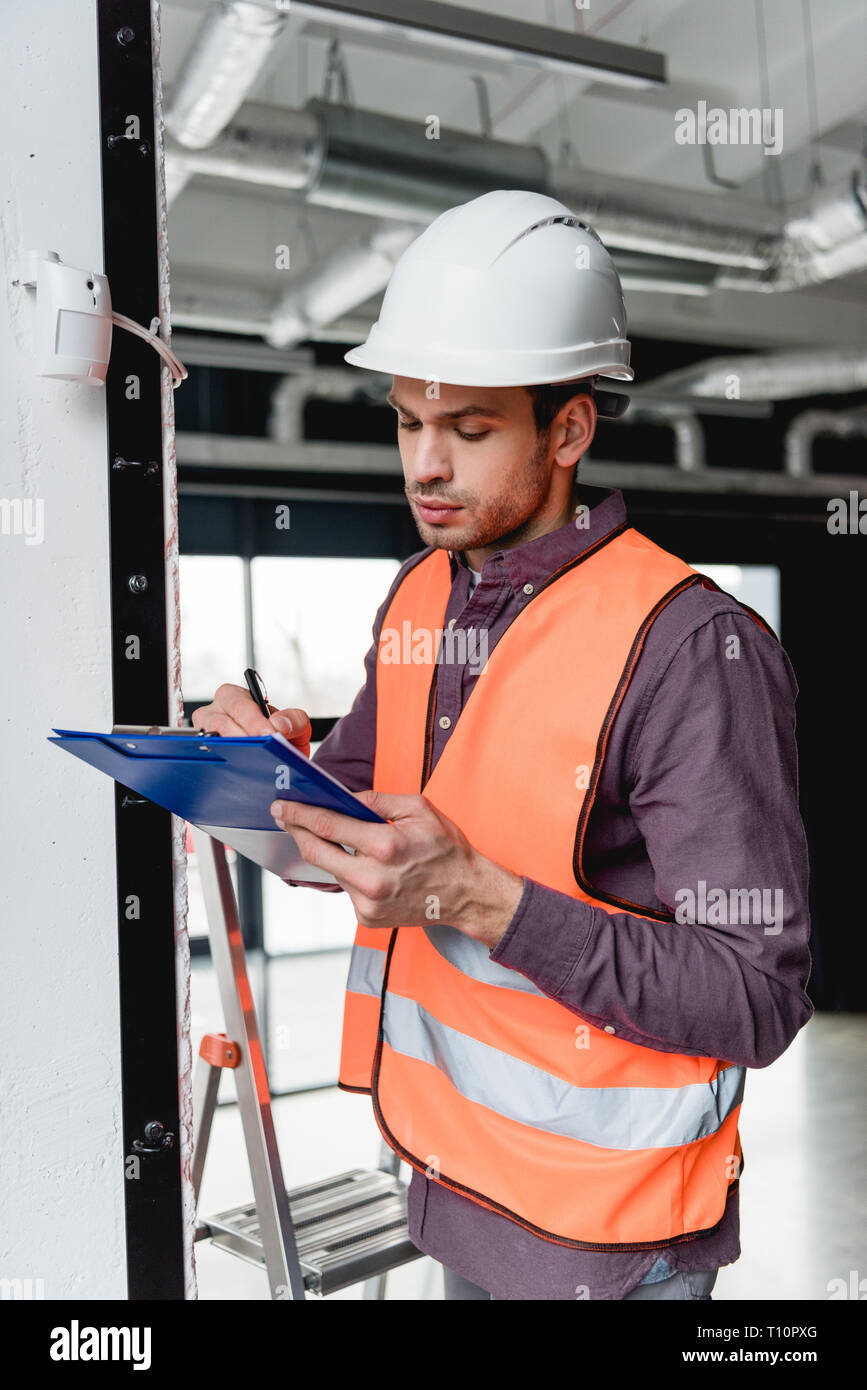 fireman in helmet standing near fire alarm while writing on clipboard ...