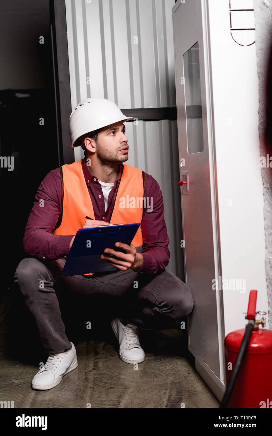thoughtful firefighter in helmet sitting and looking at fire panel ...
