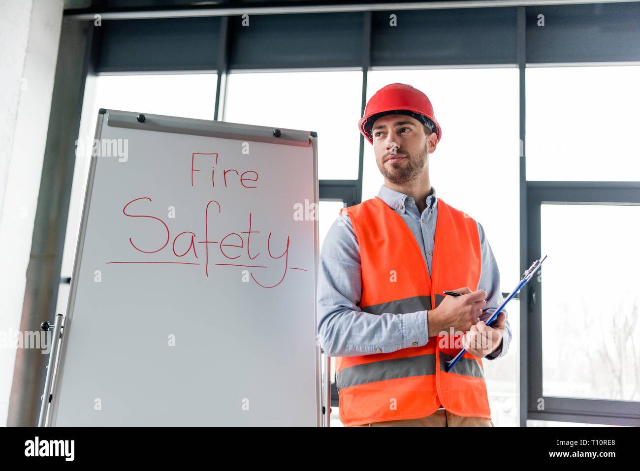 handsome firefighter in helmet holding clipboard and pen while standing ...