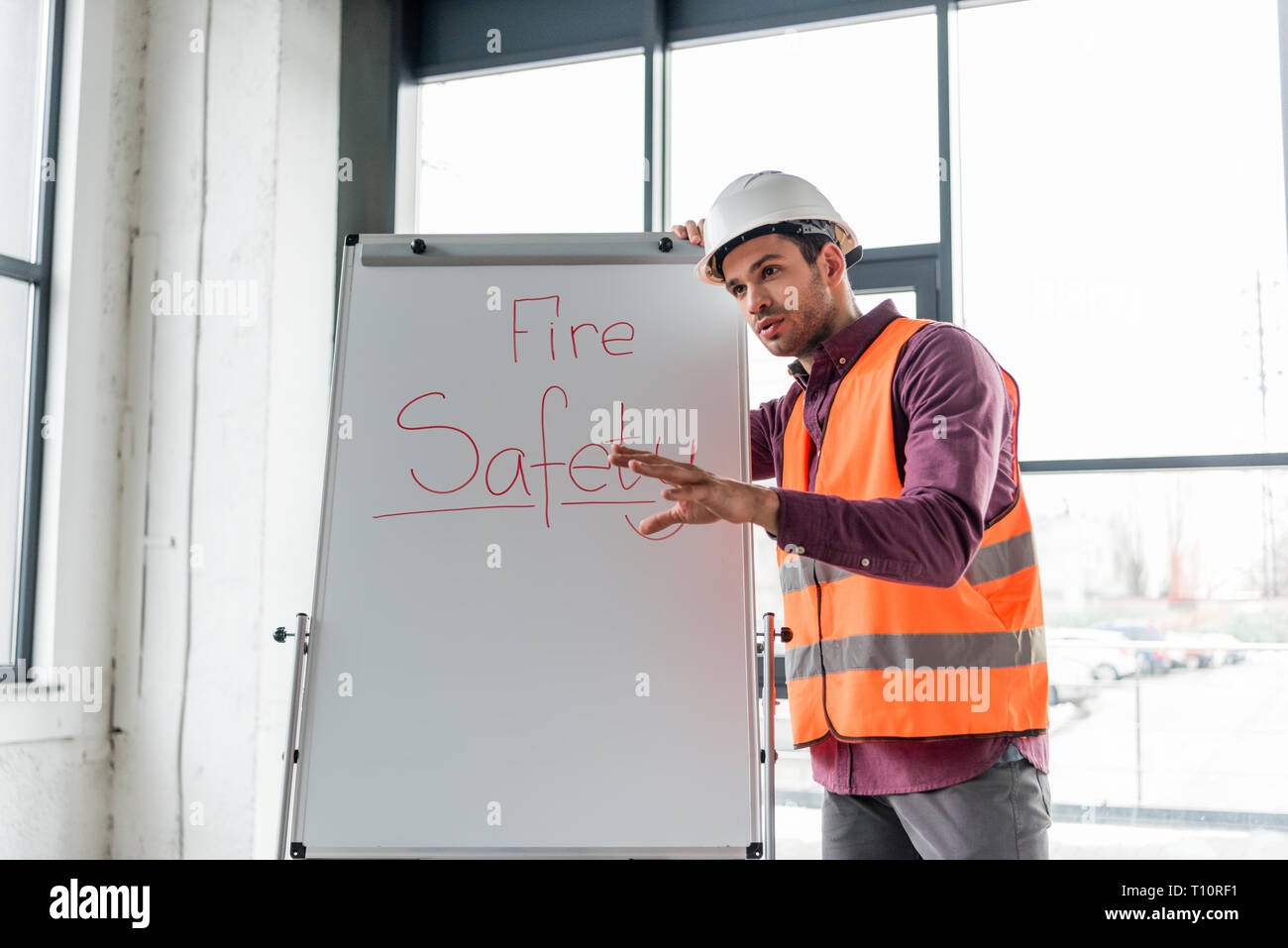 handsome firefighter in helmet talking while standing near white board ...