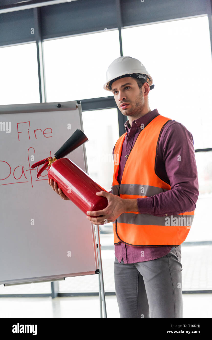 handsome fireman holding red extinguisher and standing near white board ...