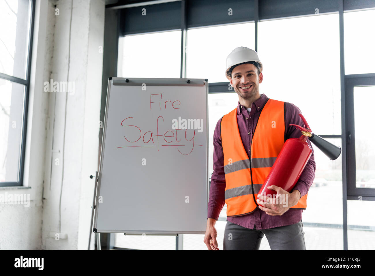 happy fireman holding red extinguisher while standing near white board ...