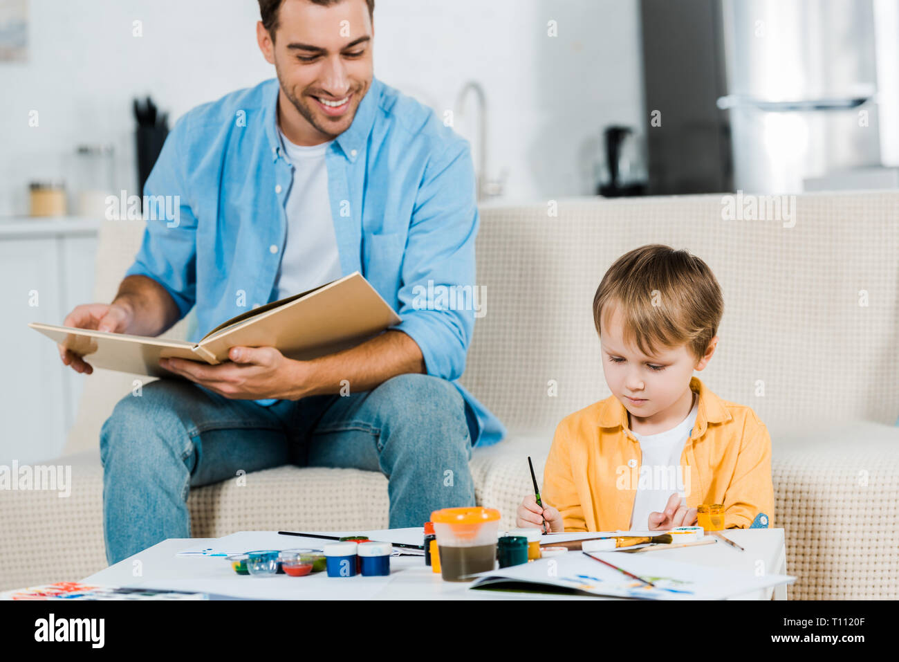 smiling father reading book while cute preschooler son drawing at home ...