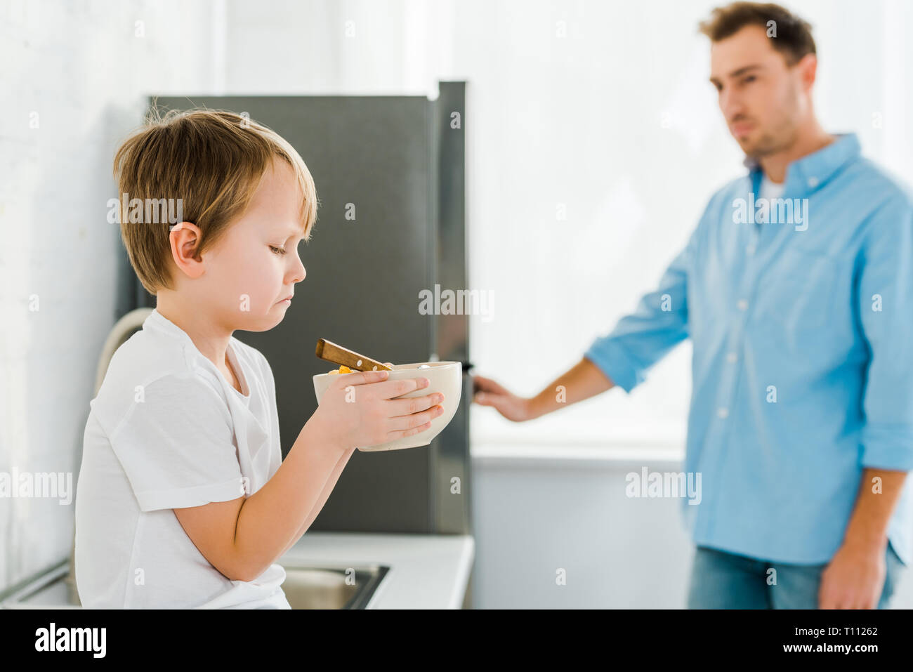 sad boy holding bowl of cereal with disappointed father on background ...