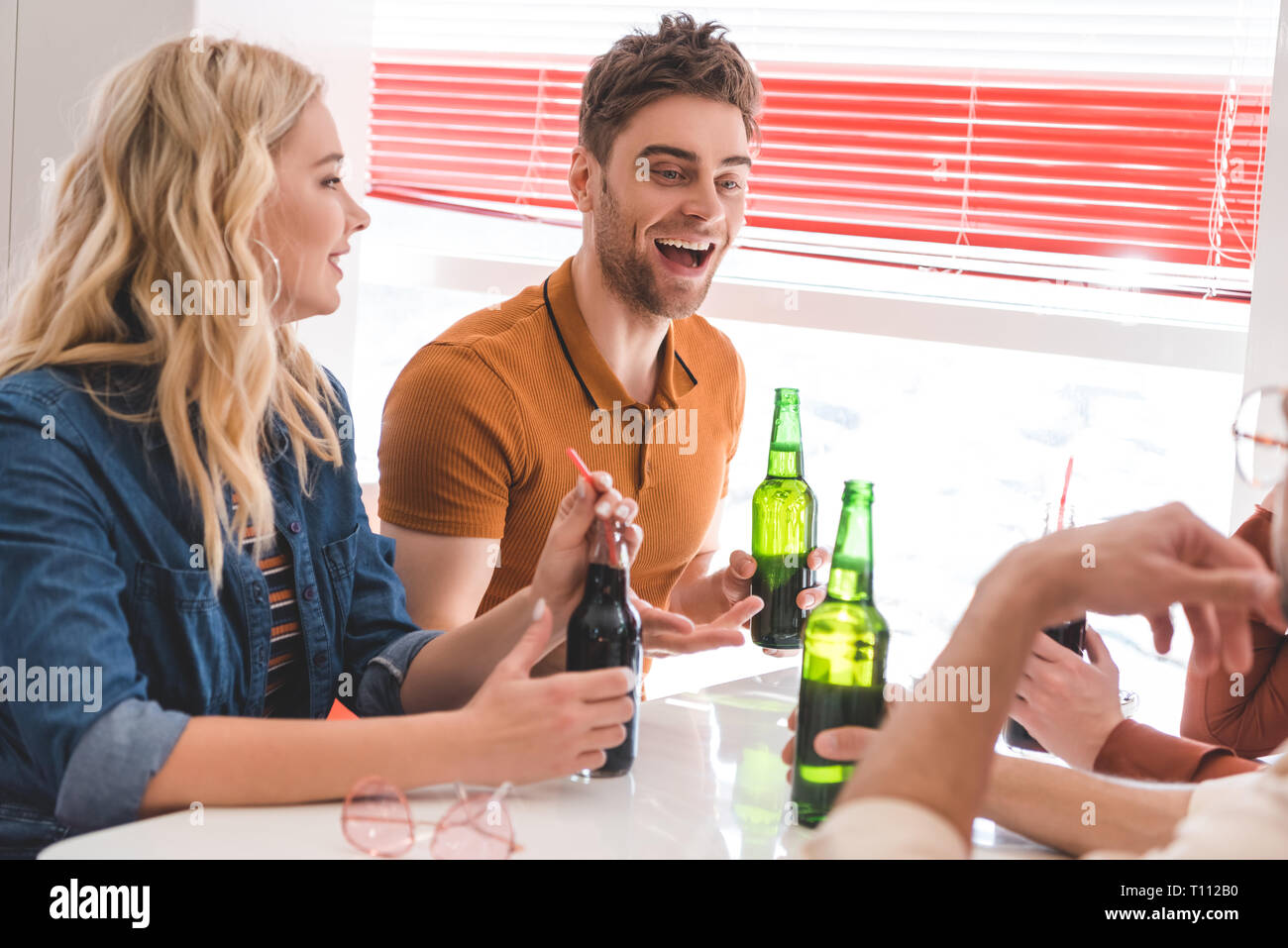 smiling friends holding glass bottles with drink and talking at cafe ...