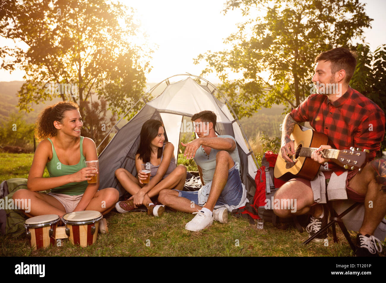 Positive young people on camping trip sitting next tent and playing ...