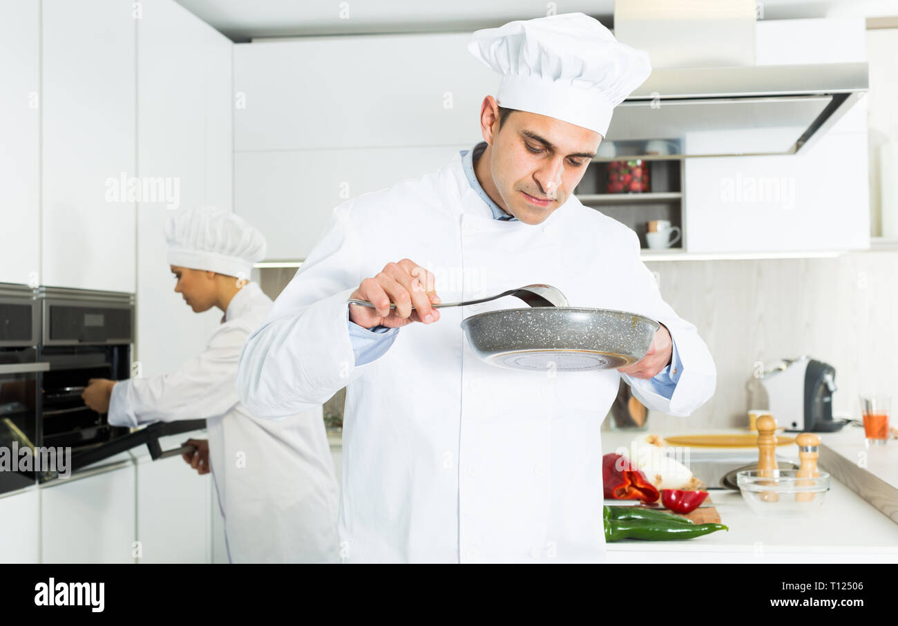 Smiling male kitchener in uniform is standing with pan in the kitchen ...