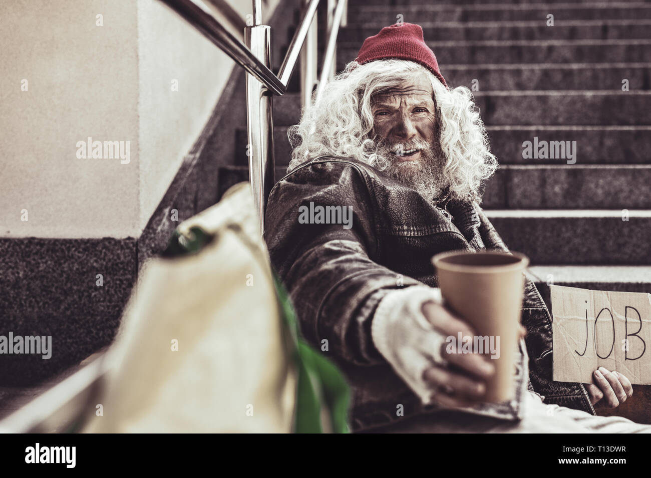 Homeless elderly offering his cap whose he holding in one hand for alms ...