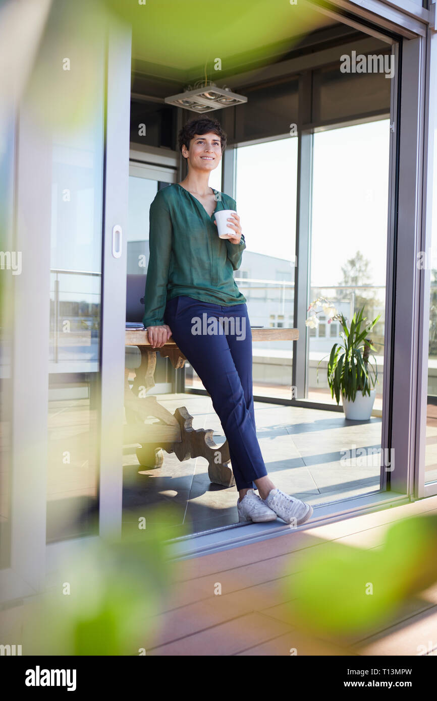 Smiling businesswoman leaning against table having a coffee break Stock ...